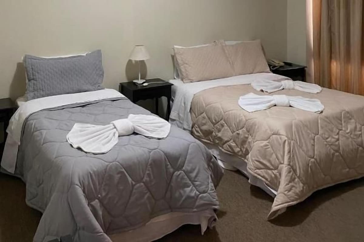 Quarto de hotel com duas camas, uma mesa de cabeceira, uma luminária e uma luz no teto.