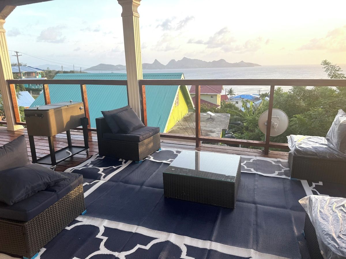Uma área de estar coberta ao ar livre, com mobiliário de vime e mesa de vidro, com vista para um corpo d'água e montanhas distantes.