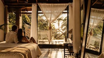 Um casal sentado na cama, de frente para uma rede e uma varanda com vista para palmeiras.