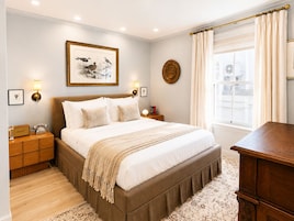 Quarto com cama grande, mesas de cabeceira, luminária na parede, quadro emoldurado e janela com cortinas.