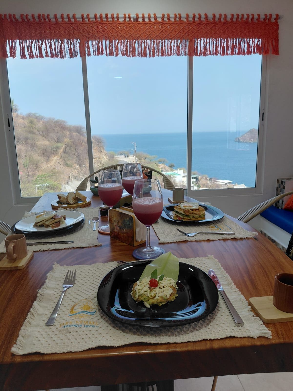 Uma mesa de jantar com pratos de comida, copos de bebida e vista para o oceano através de grandes janelas.