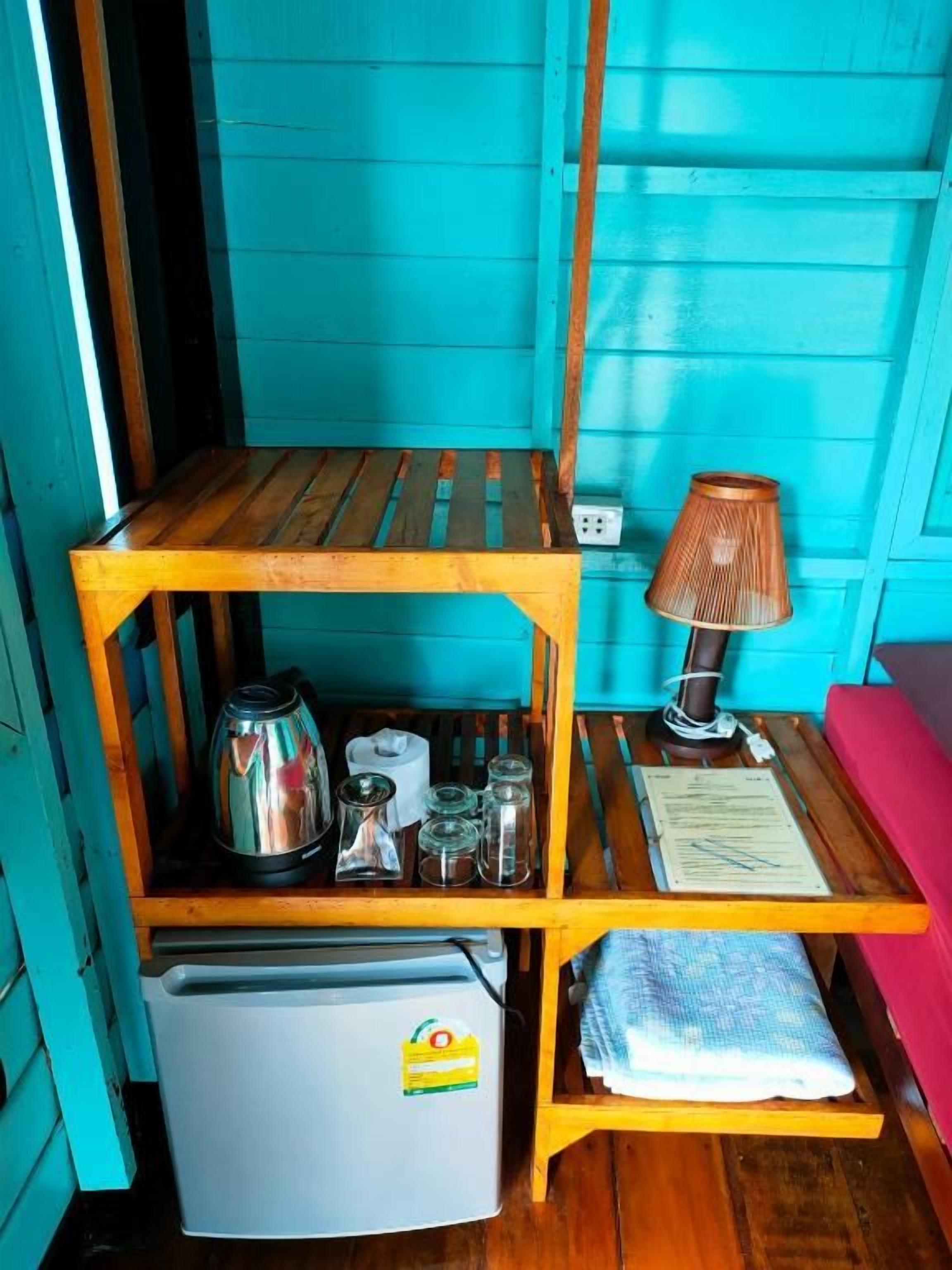 Uma kitchenette compacta com uma estante de madeira, um pequeno frigorífico, um chaleira, uma lâmpada e uma toalha.