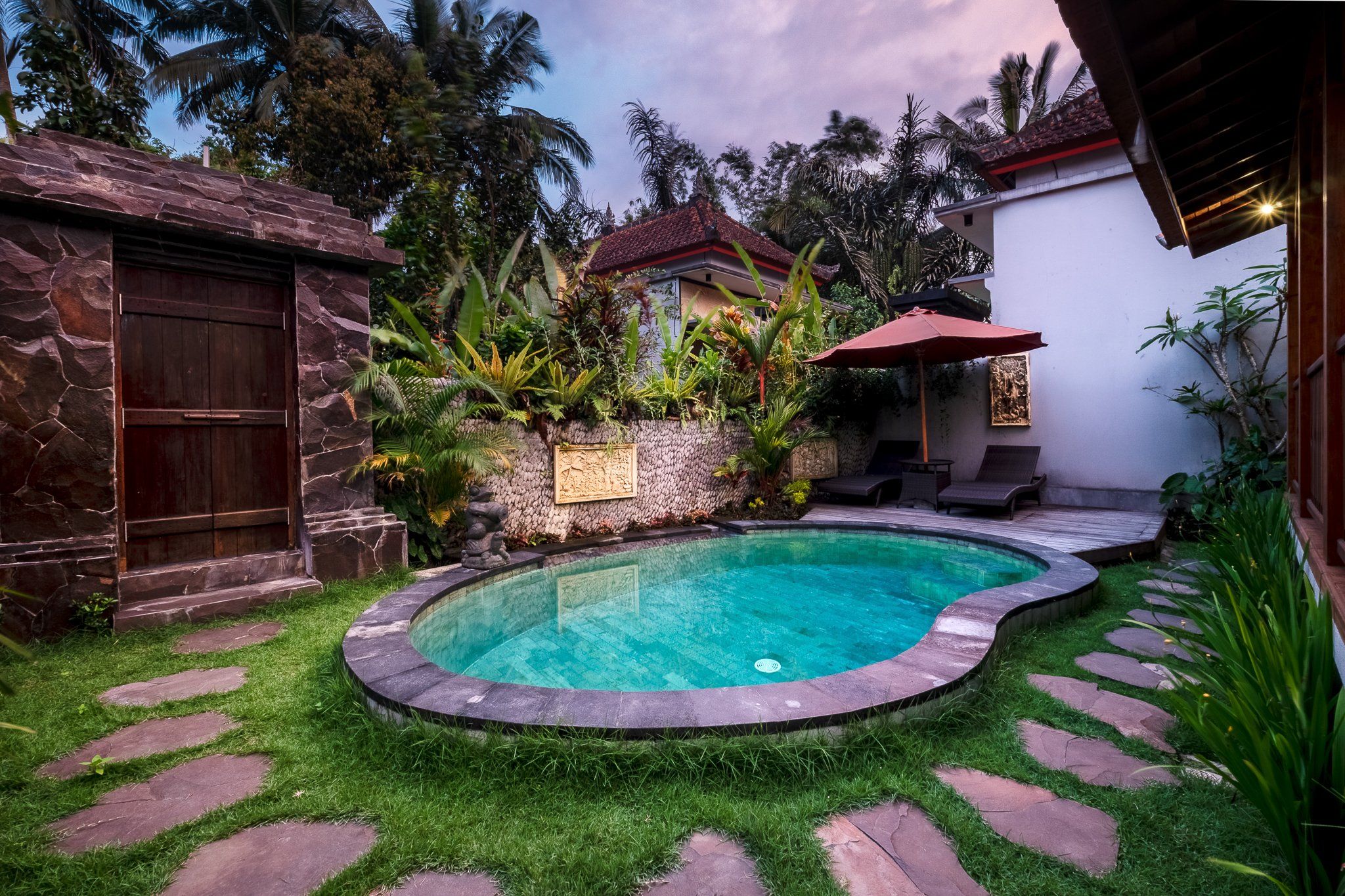 Piscina com borda de pedra, rodeada por grama e plantas, com uma parede de pedra e uma porta de madeira.