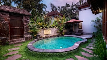 Piscina com borda de pedra, rodeada por grama e plantas, com uma parede de pedra e uma porta de madeira.