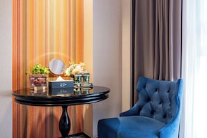 Quarto de hotel com uma mesa redonda, uma poltrona azul, um espelho e um vaso com flores.