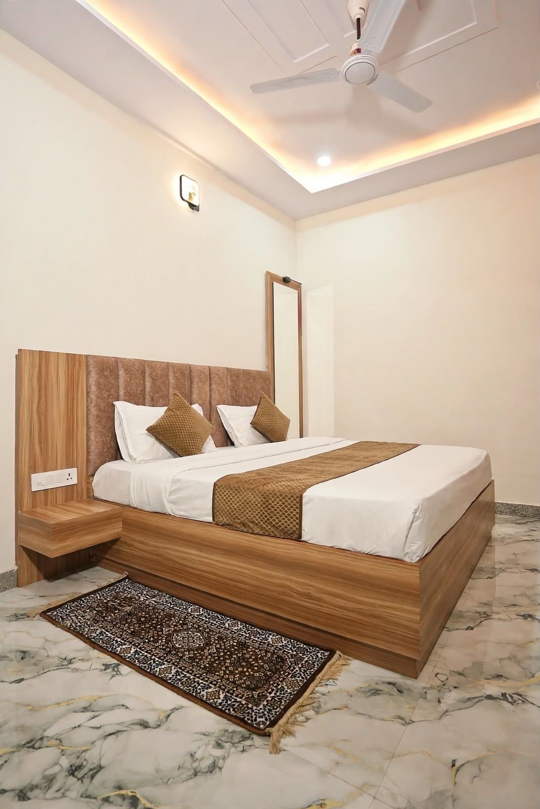 Um quarto com cama, mesa de cabeceira, espelho, ventoinha no teto e tapete estampado.