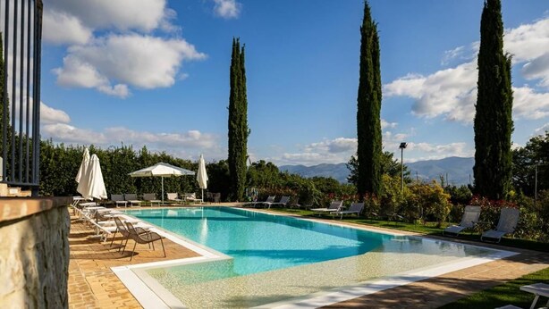 Piscina com espreguiçadeiras e guarda-sóis, cercada por árvores altas e céu claro.