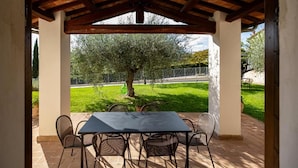 Um pátio externo com mesa e cadeiras, uma pérgola acima e vista para uma árvore e gramado.
