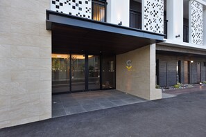 Entrada de um hotel moderno com fachada de vidro, área de acesso coberta e logotipo na parede.
