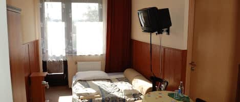 Quarto de hotel com cama, televisão na parede, uma mesinha com uma garrafa e uma janela com cortinas.
