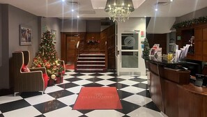 Um saguão de hotel com piso xadrez, balcão de recepção, árvore de Natal e escada que leva aos andares superiores.