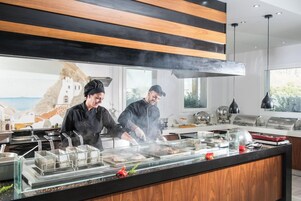 Cozinha moderna com chefs preparando comida, eletrodomésticos de aço inoxidável e vista para o mar.