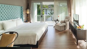 Quarto de hotel com uma cama grande, área de estar com televisão e varanda com vista para vegetação.