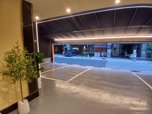 Entrada de um estacionamento com um vaso de planta, um carro e um prédio com a placa '1011' ao fundo.