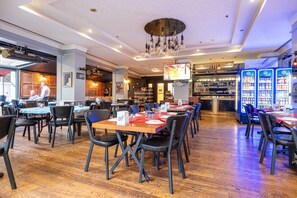 Restaurante com piso de madeira, mesas com cadeiras, área de bar e uma tela de televisão.