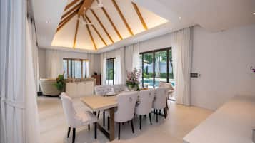 Uma sala de jantar moderna com uma mesa e cadeiras brancas, uma cozinha ao fundo e grandes janelas.