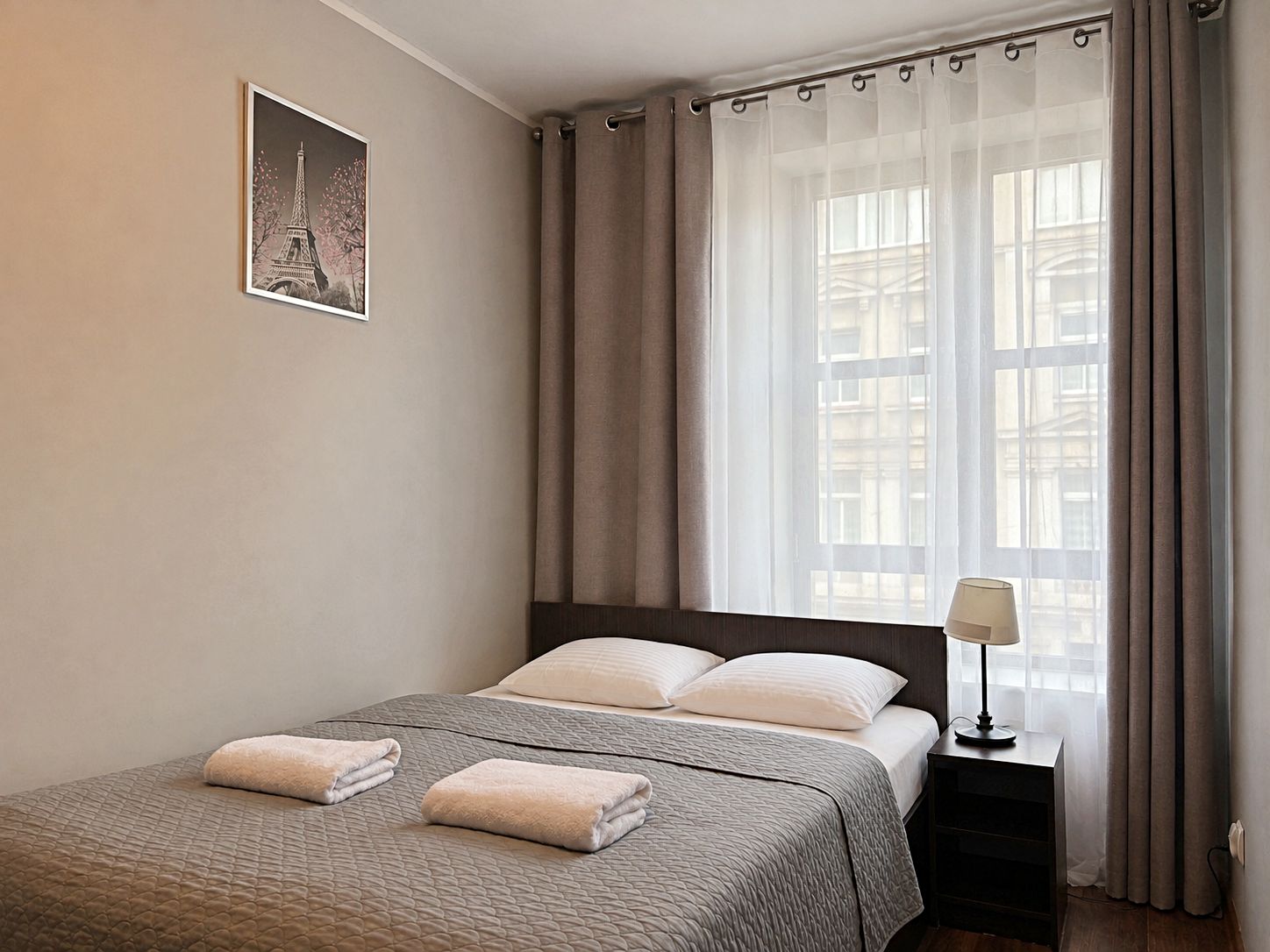 Quarto de hotel com cama, mesa de cabeceira com abajur, janela com cortinas e um quadro na parede.