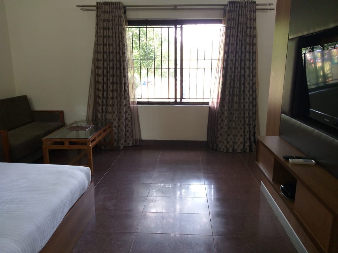 Quarto de hotel com cama, televisão, sofá, mesa pequena e janela com cortinas.