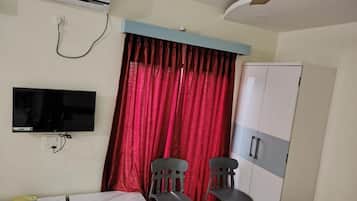 Quarto com televisão de tela plana na parede, escrivaninha com cadeira, guarda-roupa e janela com cortinas vermelhas.