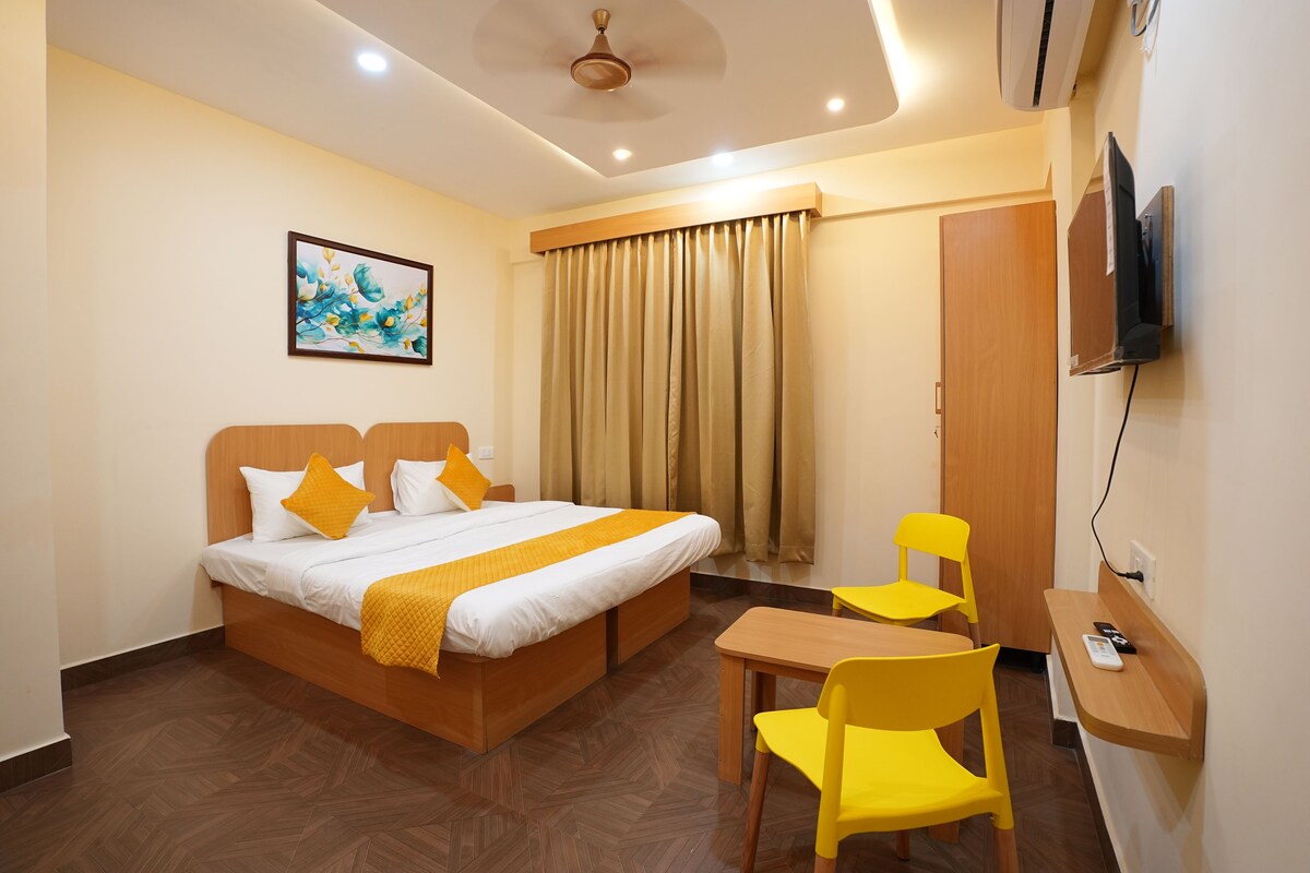 Quarto de hotel com uma cama, uma escrivaninha, duas cadeiras, uma televisão e um quadro na parede.
