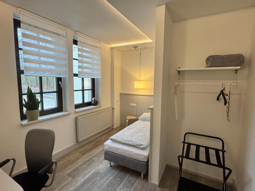 Quarto compacto com cama, mesa de cabeceira, abajur e uma cadeira.