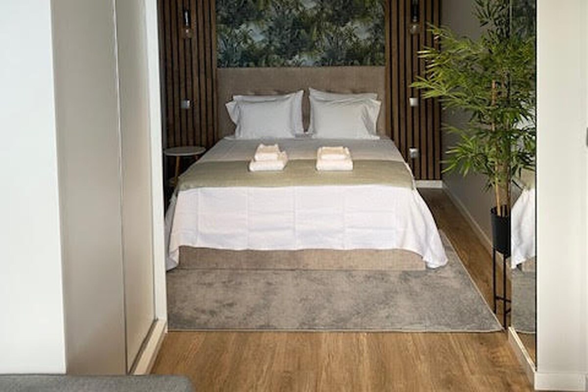 Quarto moderno com cama grande, cabeceira com ripas verticais, mesa de cabeceira, espelho e planta em vaso.
