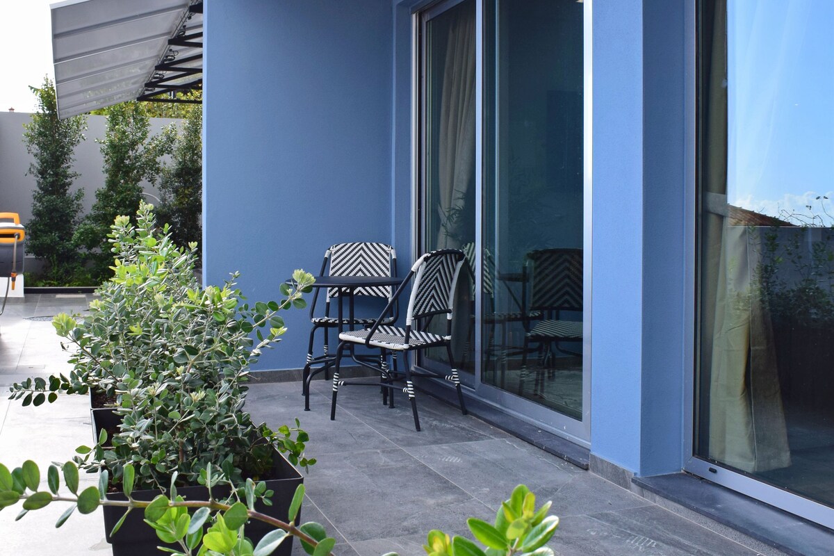 Uma área de estar moderna ao ar livre, com parede azul, grandes portas de vidro deslizantes e plantas em vasos.