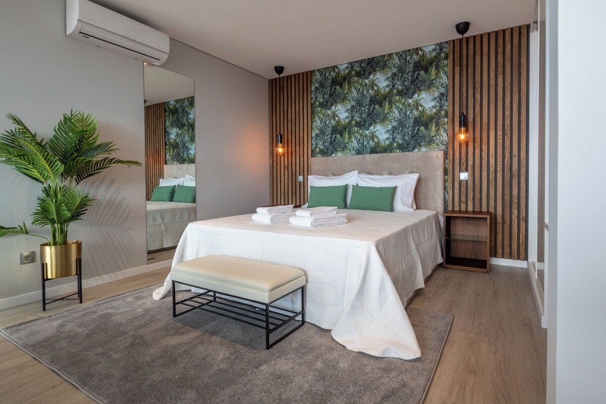 Quarto moderno com uma cama grande, mesa de cabeceira, um banco, planta em vaso e ar condicionado fixado na parede.