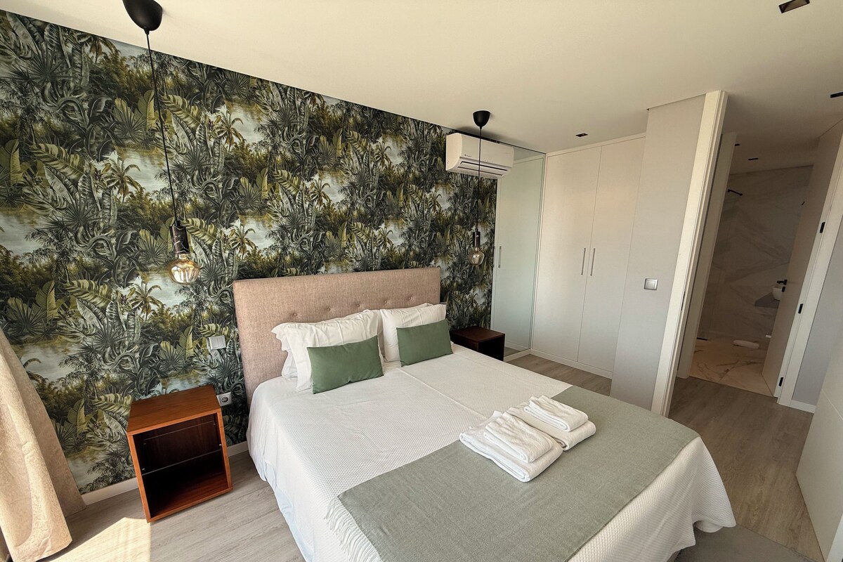 Quarto com cama grande, mesa de cabeceira, guarda-roupa e parede com mural tropical.