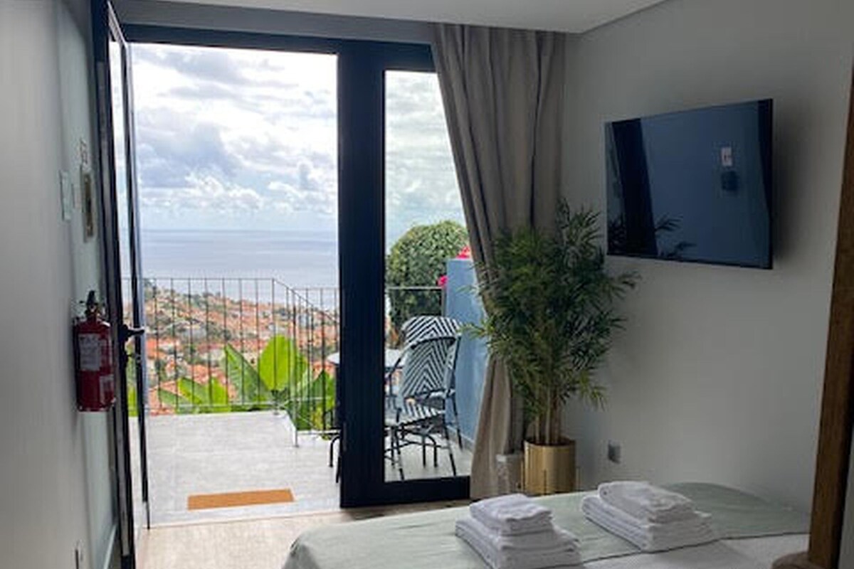 Quarto de hotel com cama, televisão fixada na parede, planta em vaso e vista para o mar através de uma porta aberta.