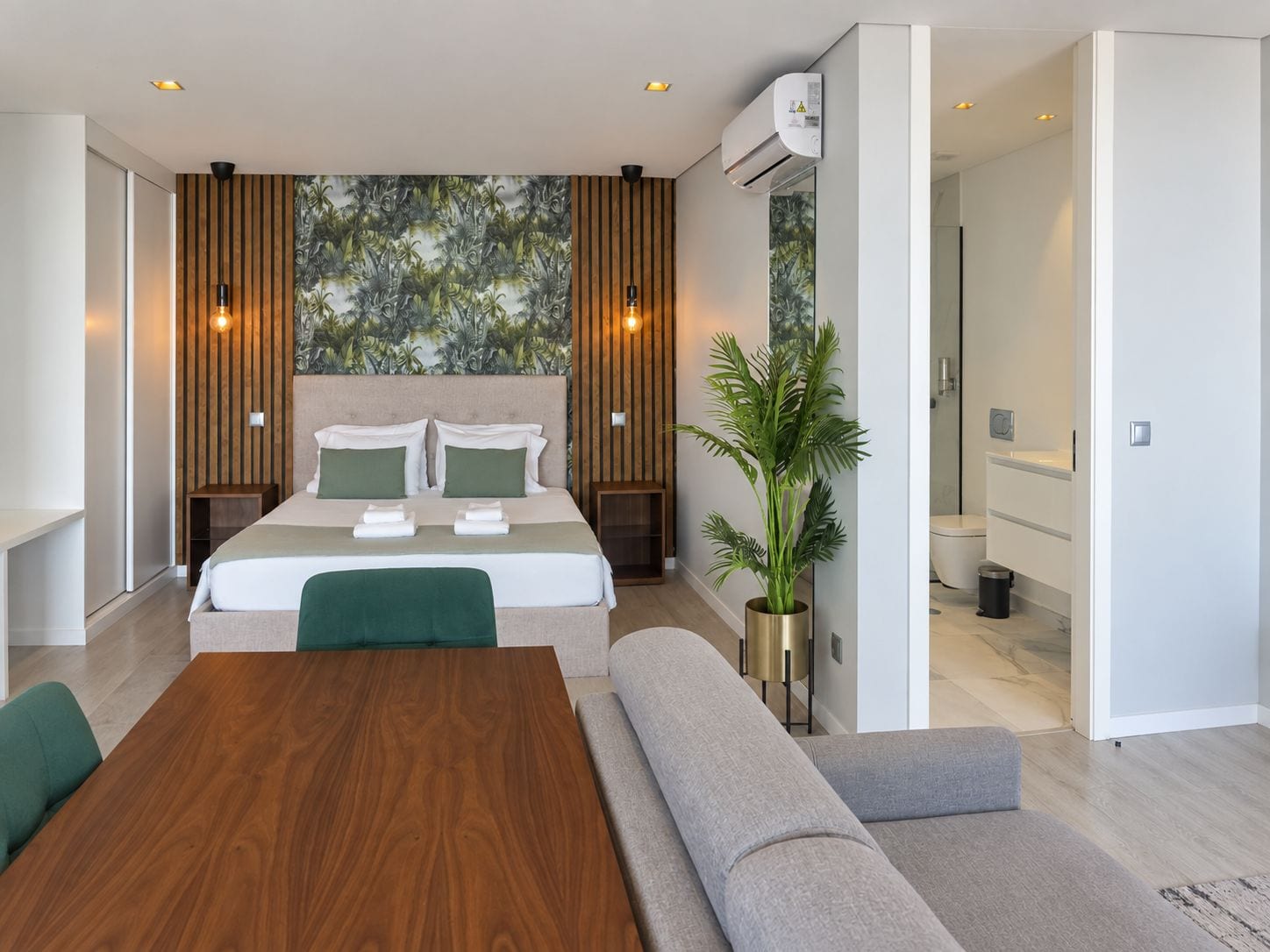 Quarto de hotel moderno com uma cama grande, uma mesa de jantar de madeira, um sofá cinza e um vaso com planta.