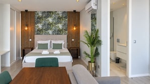 Quarto de hotel moderno com uma cama grande, uma mesa de jantar de madeira, um sofá cinza e um vaso com planta.