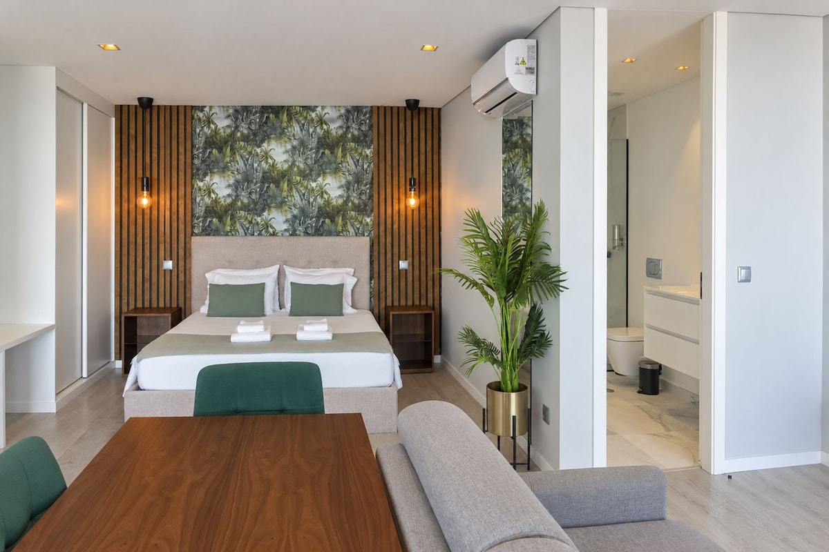Quarto de hotel moderno com uma cama grande, uma mesa de jantar de madeira, um sofá cinza e um vaso com planta.