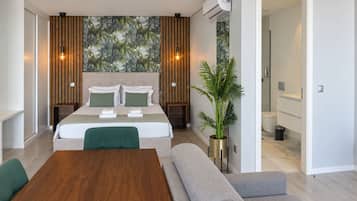 Quarto de hotel moderno com uma cama grande, uma mesa de jantar de madeira, um sofá cinza e um vaso com planta.