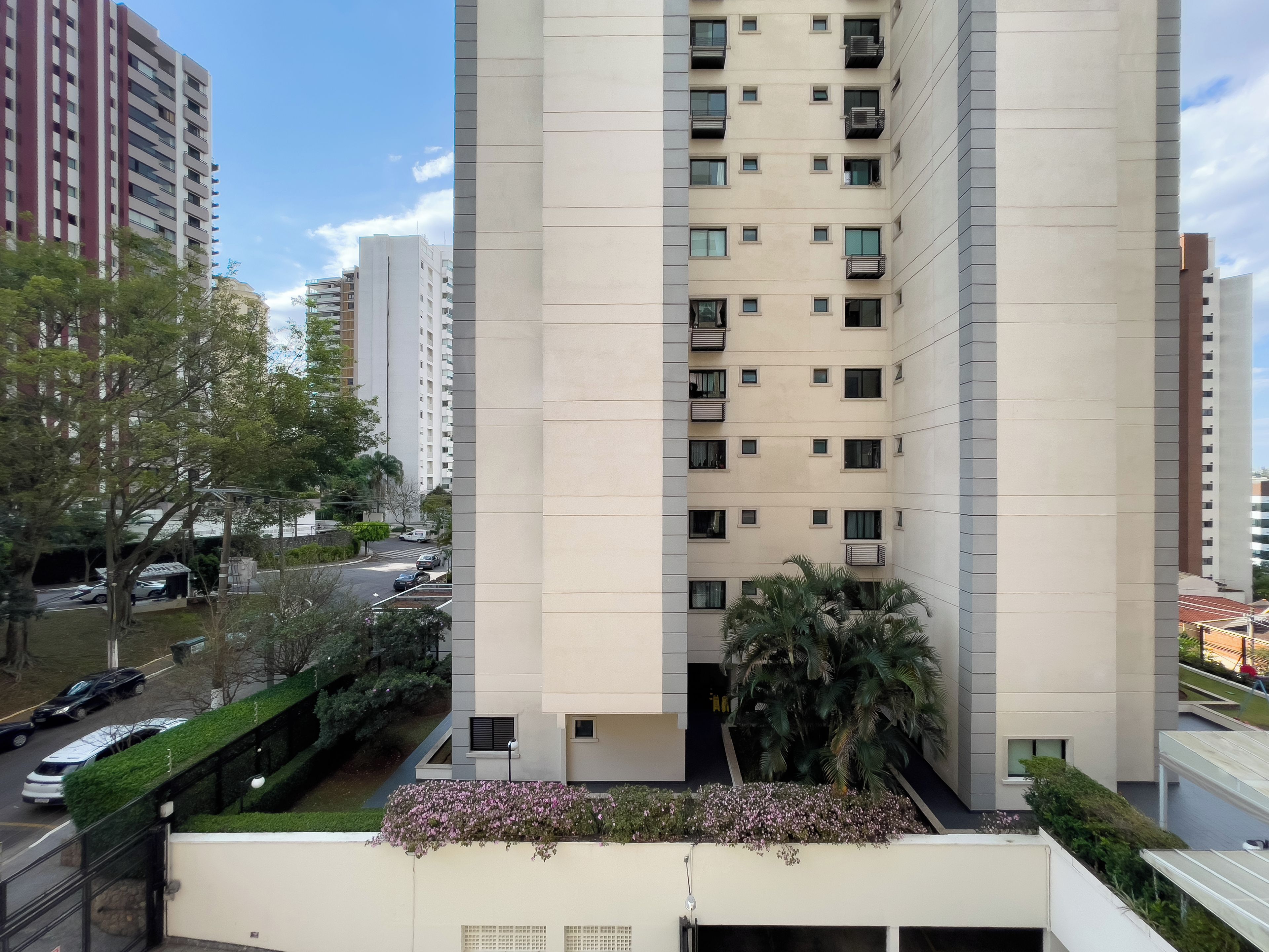 Edifício residencial com vários andares, varandas, área de estacionamento e um pequeno jardim com flores.