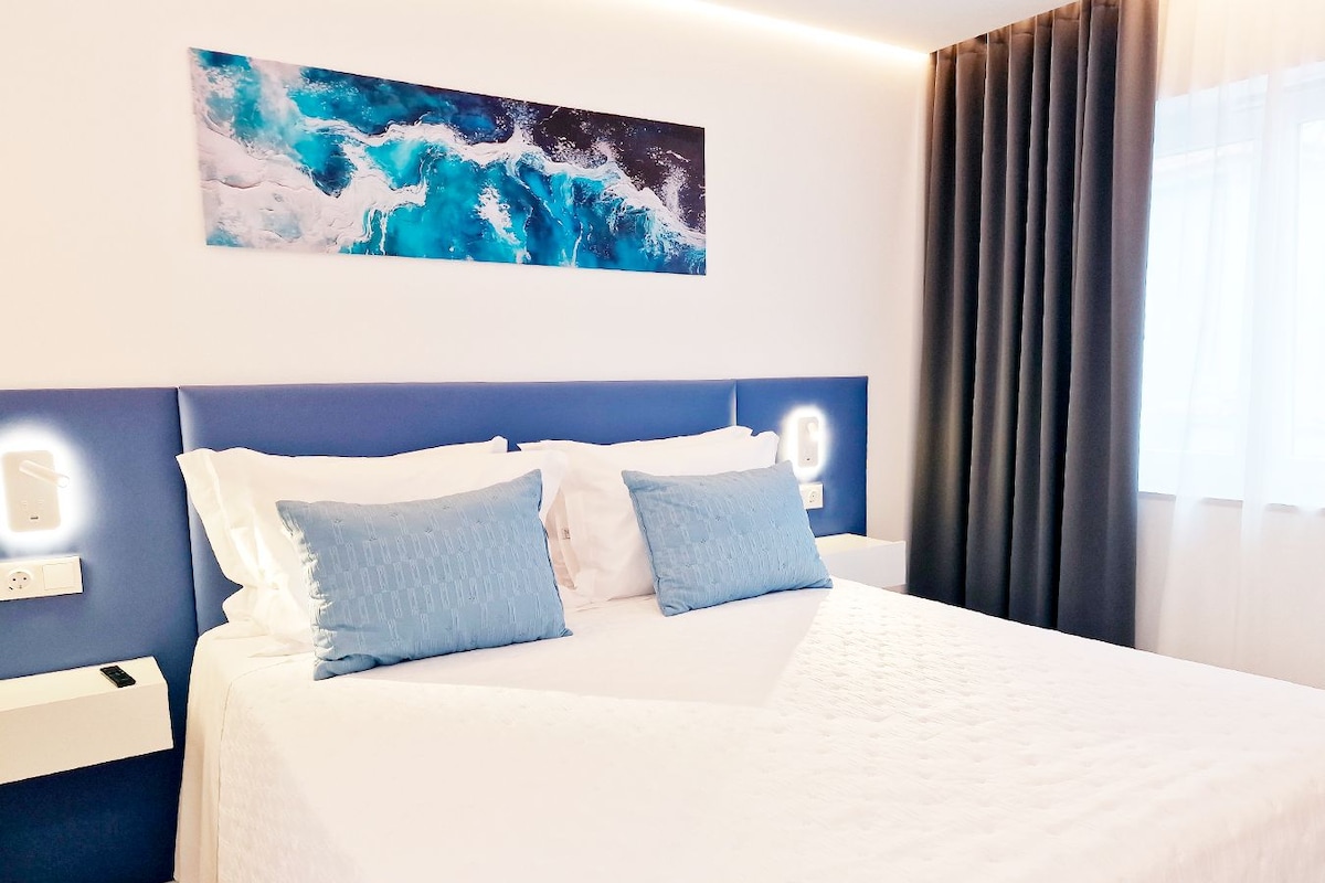 Quarto de hotel com uma cama grande, cabeceira, luminárias laterais, mesa de cabeceira e um quadro na parede.