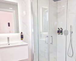 Casa de banho moderna com chuveiro em cabine de vidro, lavatório branco com espelho e artigos de higiene pessoal na parede.