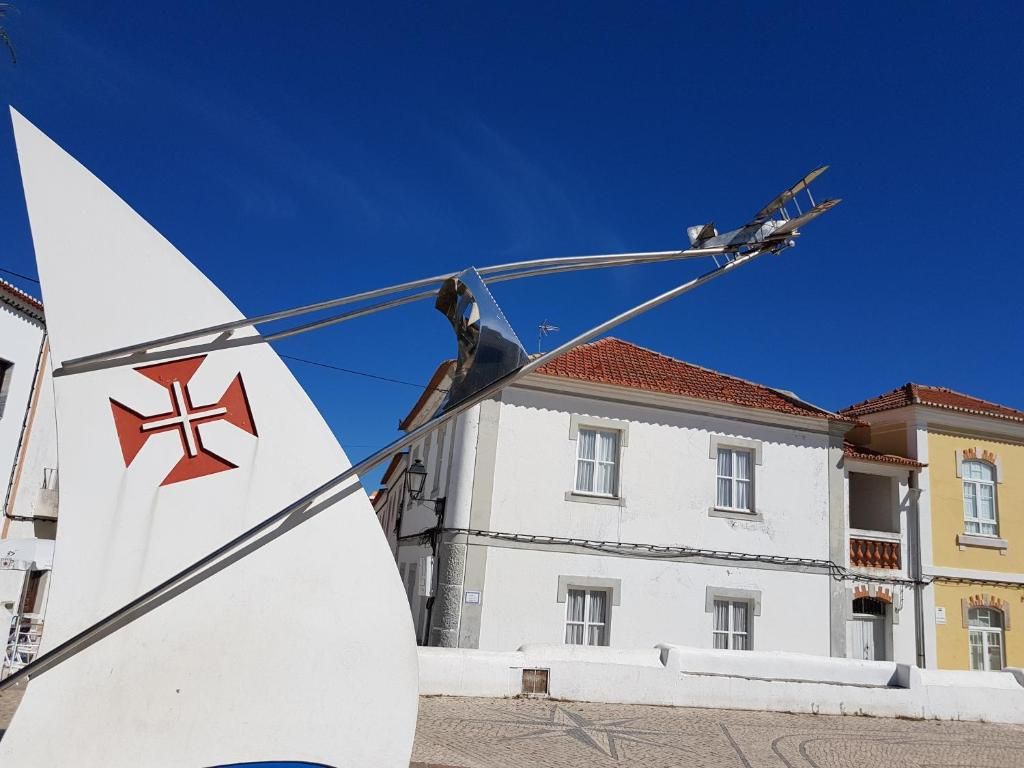 Um veleiro com o emblema de uma cruz vermelha, atracado perto de edifícios com telhado em tijolos vermelhos.
