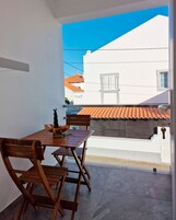 Varanda com mesa e cadeiras de madeira, vista para um prédio branco com telhas laranja.