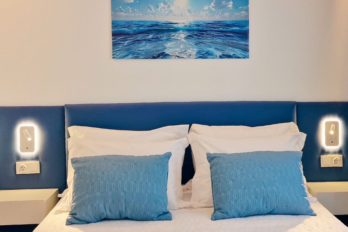 Quarto com cama, duas almofadas, um quadro pendurado na parede com vista para o oceano e luminárias de cabeceira.