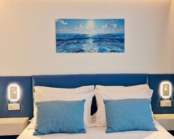 Quarto com cama, duas almofadas, um quadro pendurado na parede com vista para o oceano e luminárias de cabeceira.