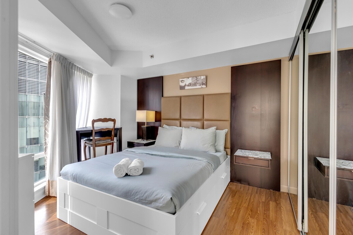 Quarto de hotel moderno com cama grande, escrivaninha, cadeira, mesa de cabeceira e janela com cortinas.