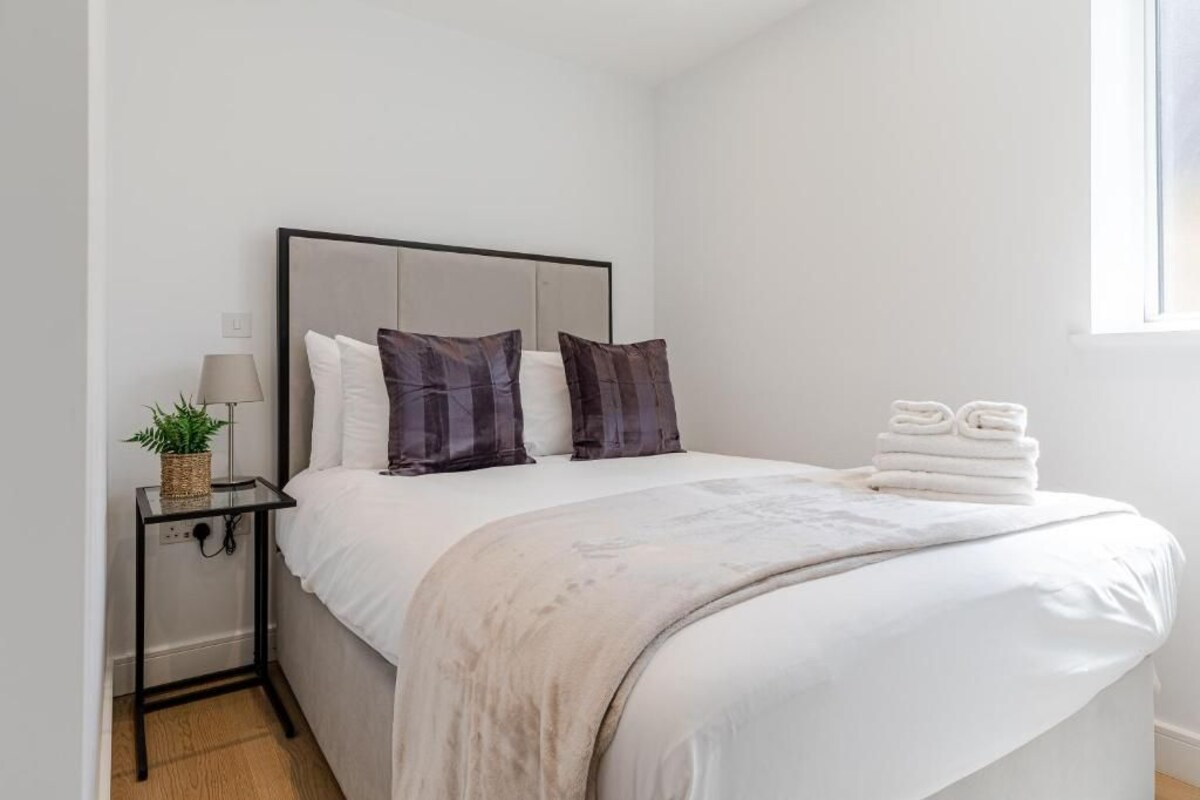 Cama bem arrumada com cabeceira, mesa de cabeceira com abajur e uma janela.