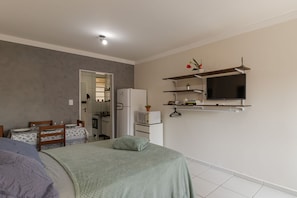 Quarto compacto com cama, escrivaninha e televisão na parede.