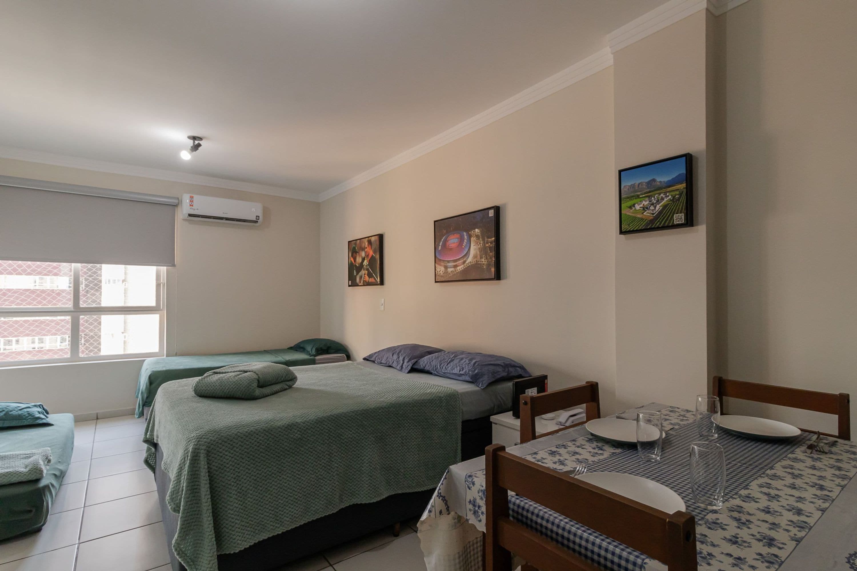 Quarto com duas camas, uma mesa de jantar e cadeiras, e uma janela com vista para edifícios.