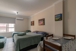 Quarto com duas camas, uma mesa de jantar e cadeiras, e uma janela com vista para edifícios.