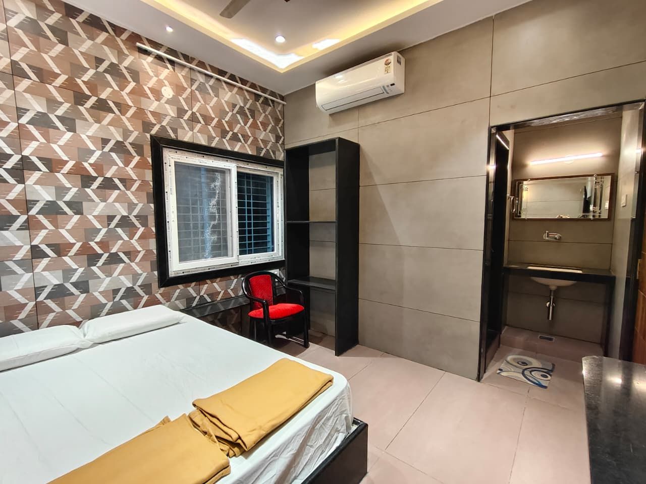 Quarto de hotel com cama, uma cadeira vermelha, janela, ar condicionado fixado na parede e banheiro com pia e espelho.