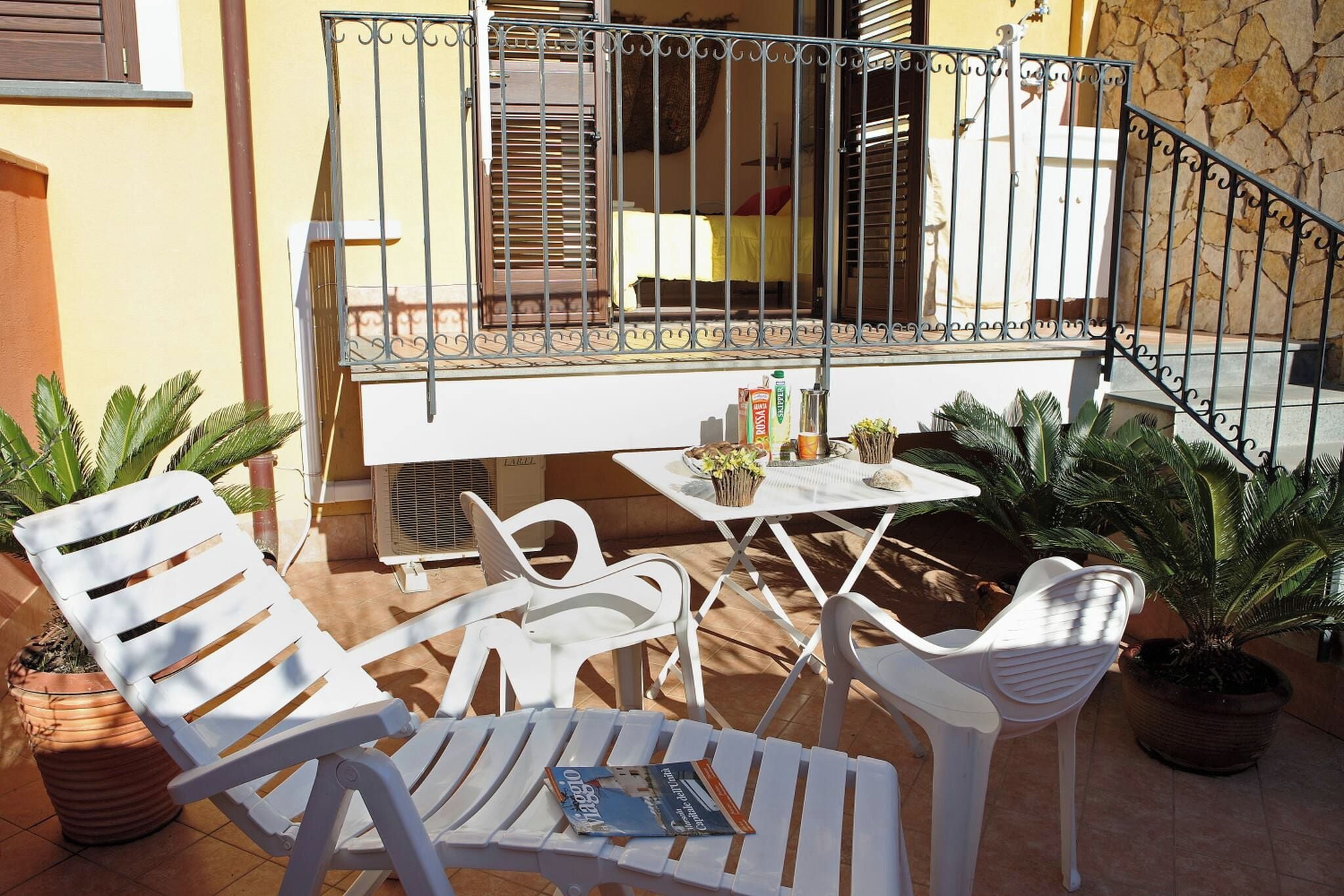 Varanda com cadeiras de plástico branco, uma mesa e um pequeno vaso de planta, além de corrimão de metal.