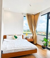 Quarto de hotel moderno com uma cama grande, mesa de cabeceira, planta em vaso e vista da cidade através de uma janela ampla.