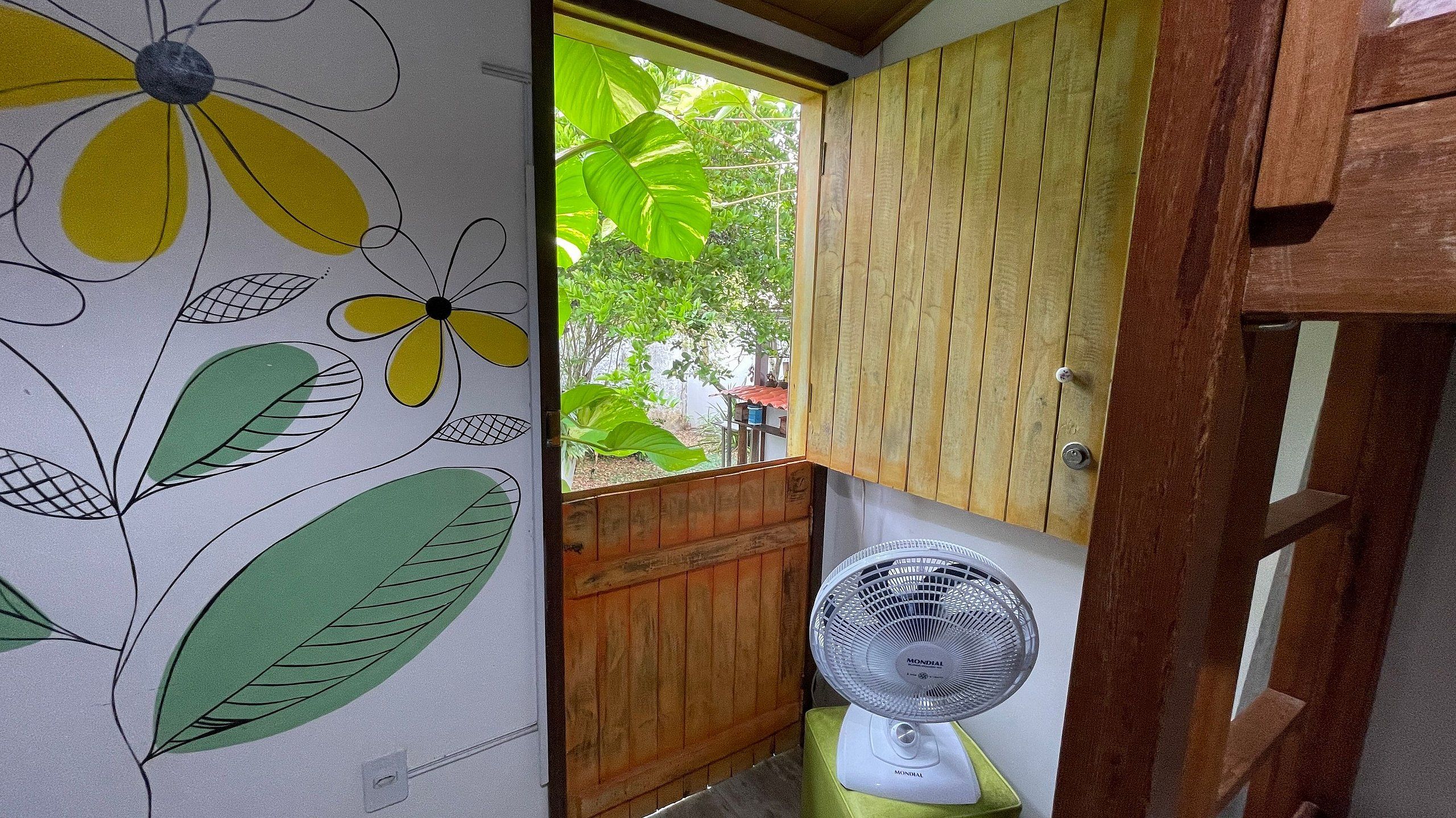 Quarto com porta de madeira, um ventilador e janela com vista para o verde.